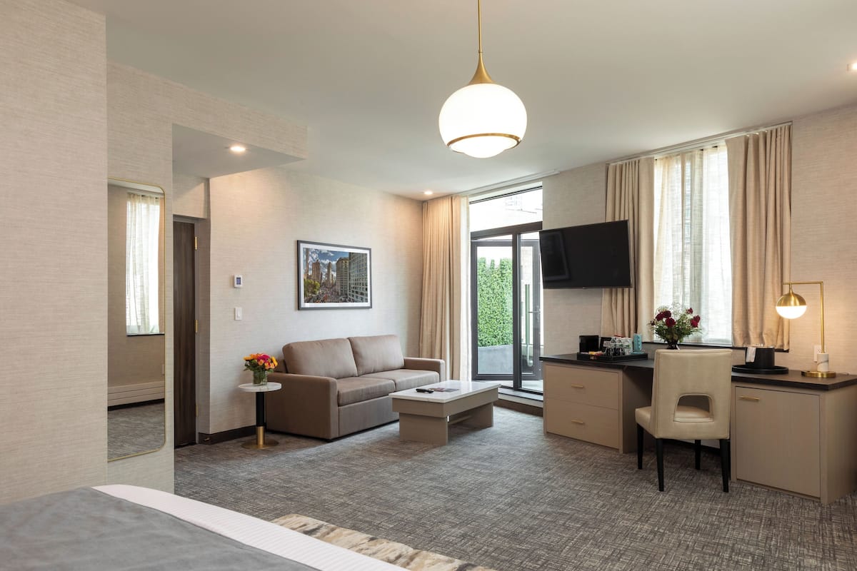 Quarto de hotel moderno com sofá, mesa de centro, escrivaninha e cadeira. Há uma TV na parede, uma janela com cortinas e uma luminária no teto.