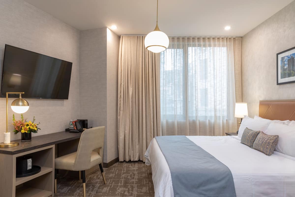 Quarto de hotel com cama, escrivaninha, cadeira, televisão e janela com cortinas.