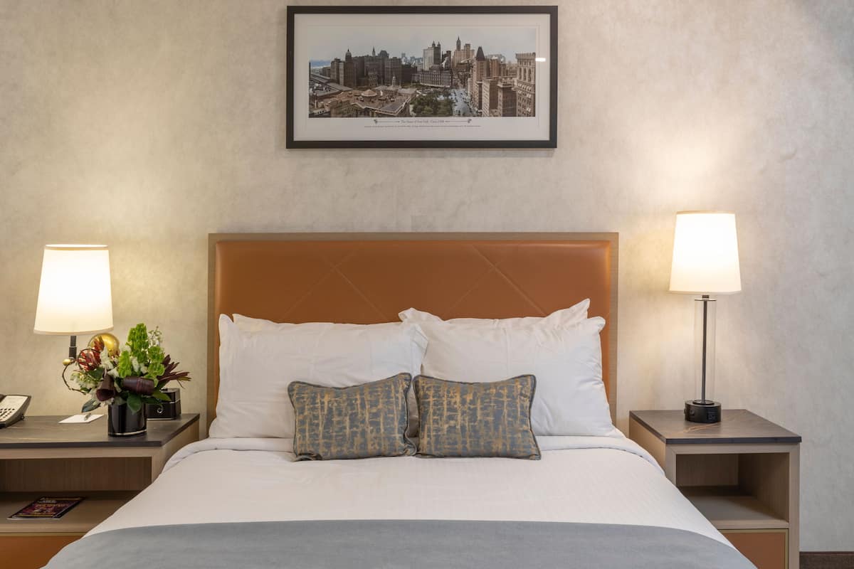 Quarto de hotel com uma cama grande, duas mesas de cabeceira, um quadro com vista urbana e uma mesa de apoio com abajur.