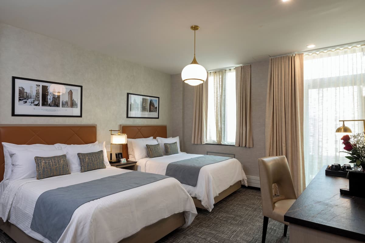 Quarto de hotel com duas camas, uma escrivaninha e uma cadeira. Há quadros nas paredes, uma janela com cortinas e um lustre.