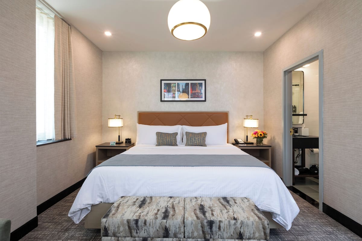 Quarto de hotel com cama grande, duas mesas de cabeceira com luminárias, cabeceira e um quadro emoldurado na parede, além de um espelho no canto.