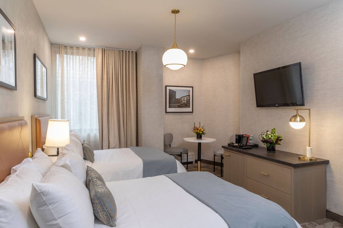 Quarto de hotel com duas camas, uma escrivaninha, televisão e janela com cortinas.
