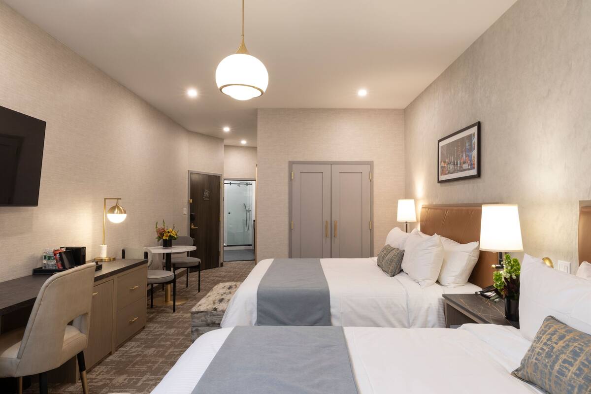 Quarto de hotel com duas camas, uma escrivaninha, uma cadeira, uma mesinha e zona de chuveiro.