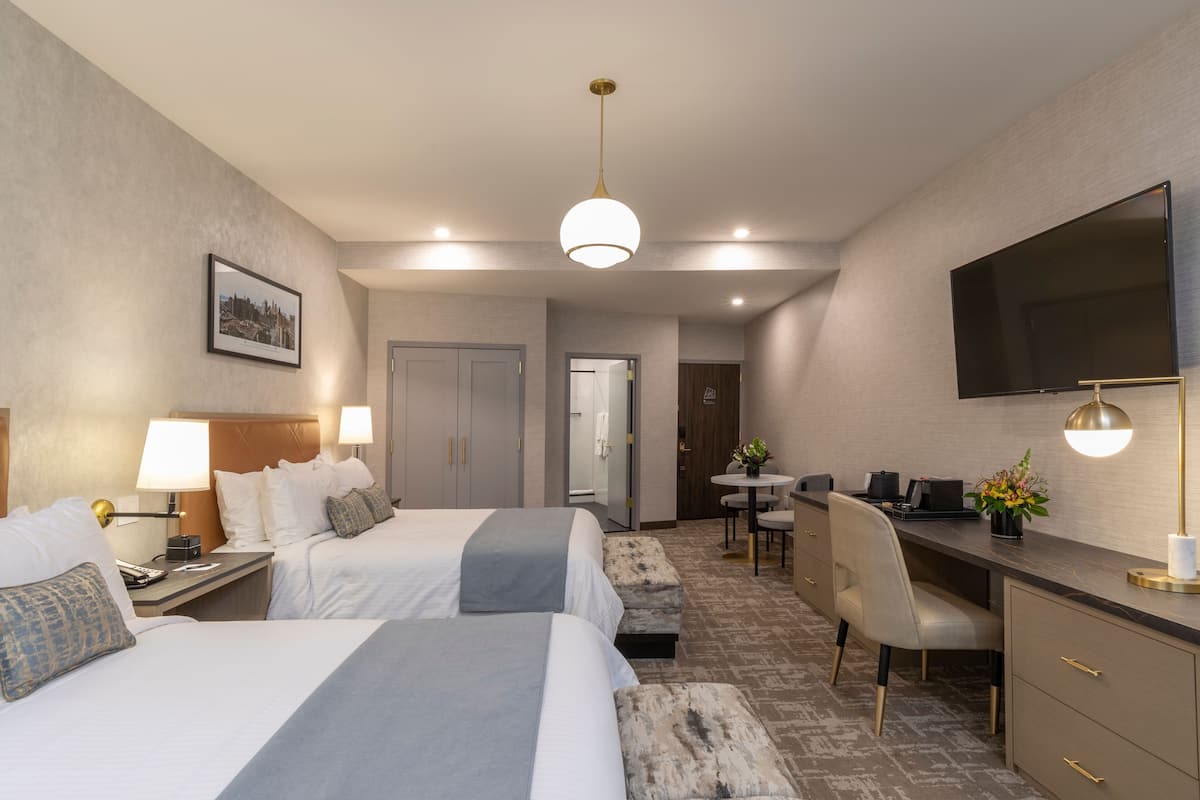 Quarto de hotel com duas camas, uma escrivaninha, uma cadeira, uma televisão e uma pequena mesa com um vaso de flores.
