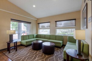 Quarto de hotel com sofá modular verde, dois pufes redondos, uma mesinha de centro com abajur e janelas amplas com persianas.