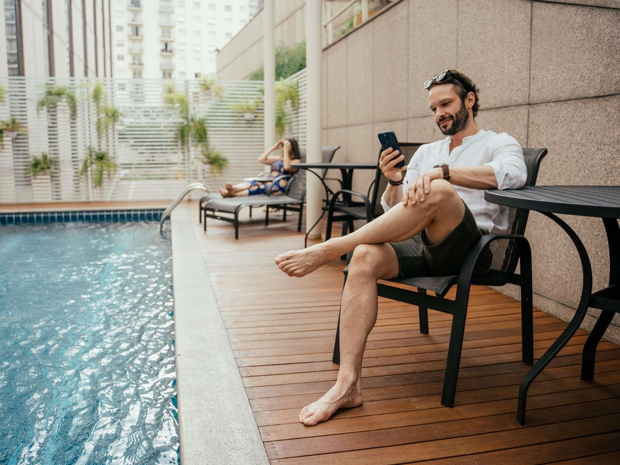 Um homem sentado em uma espreguiçadeira à beira da piscina, segurando um smartphone.