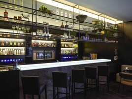 Uma área de bar com várias prateleiras de garrafas de bebidas, um balcão e diversos bancos.