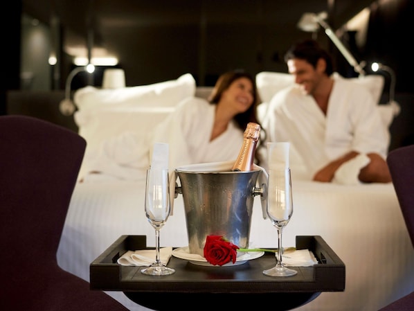 Uma cama com roupa de cama branca, uma mesa de cabeceira com um tabuleiro que contém uma garrafa de champanhe, dois copos e uma rosa vermelha.