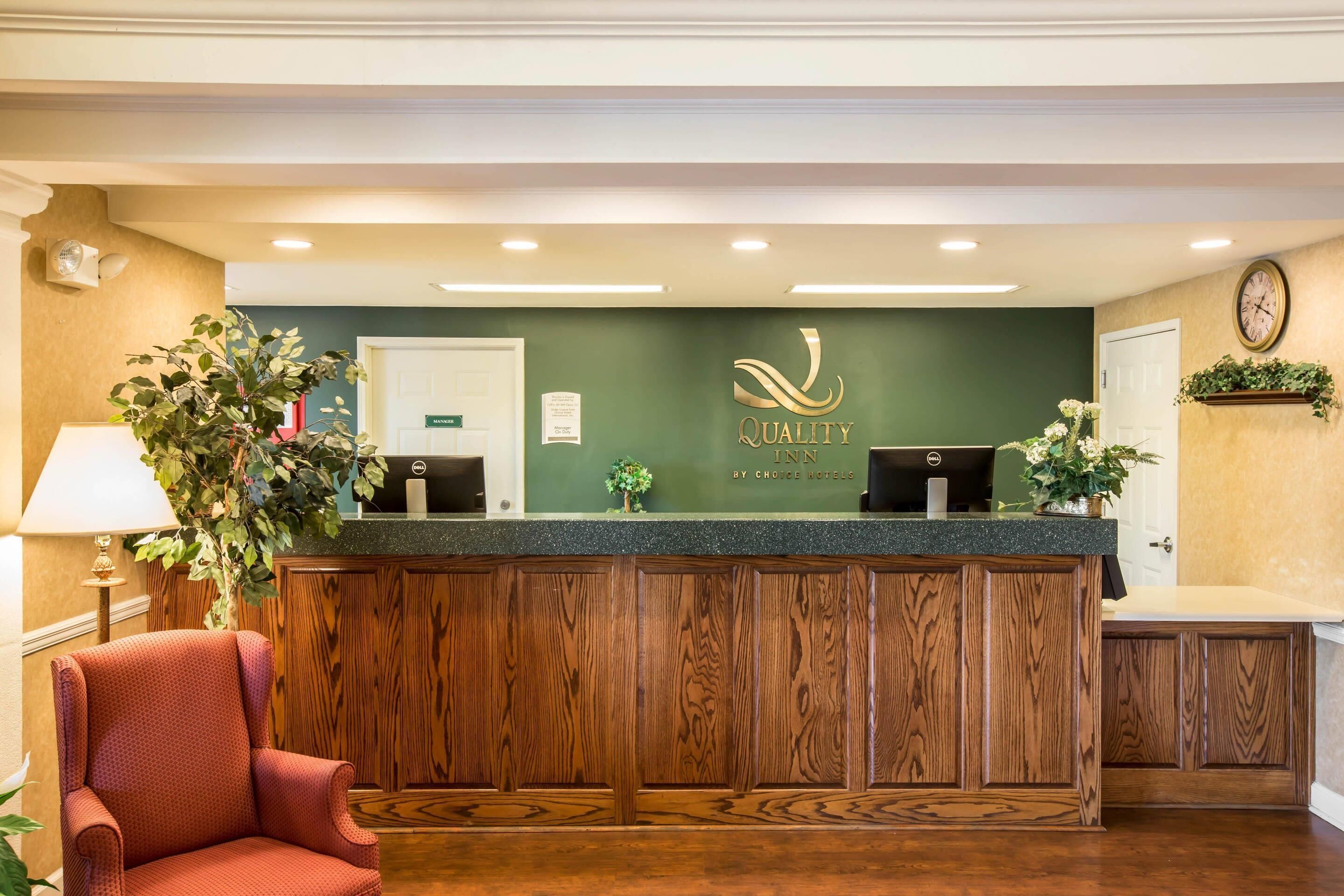 Uma receção com design em painel de madeira, uma parede verde com o logótipo 'Quality Inn', e uma área de receção com uma cadeira e uma planta.