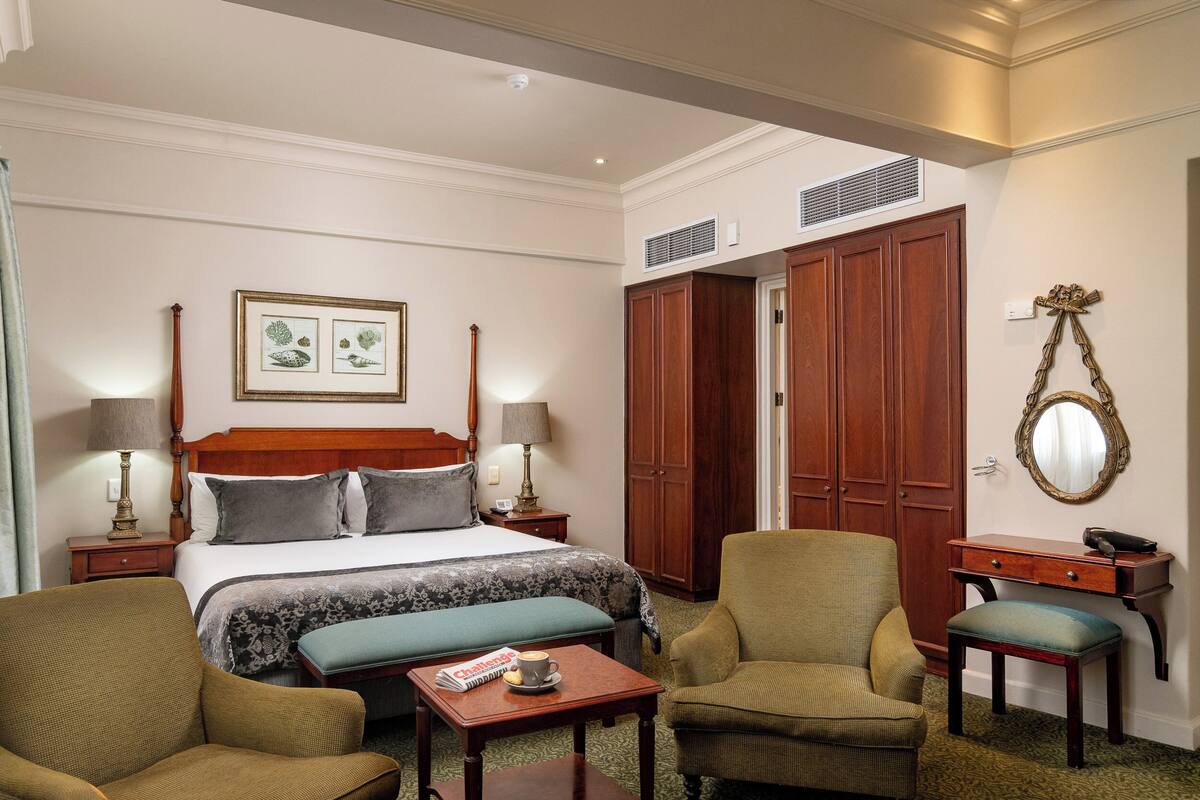 Quarto de hotel com uma cama grande, duas mesas de cabeceira com abajures, um guarda-roupa de madeira, uma pequena escrivaninha com cadeira e um espelho na parede.