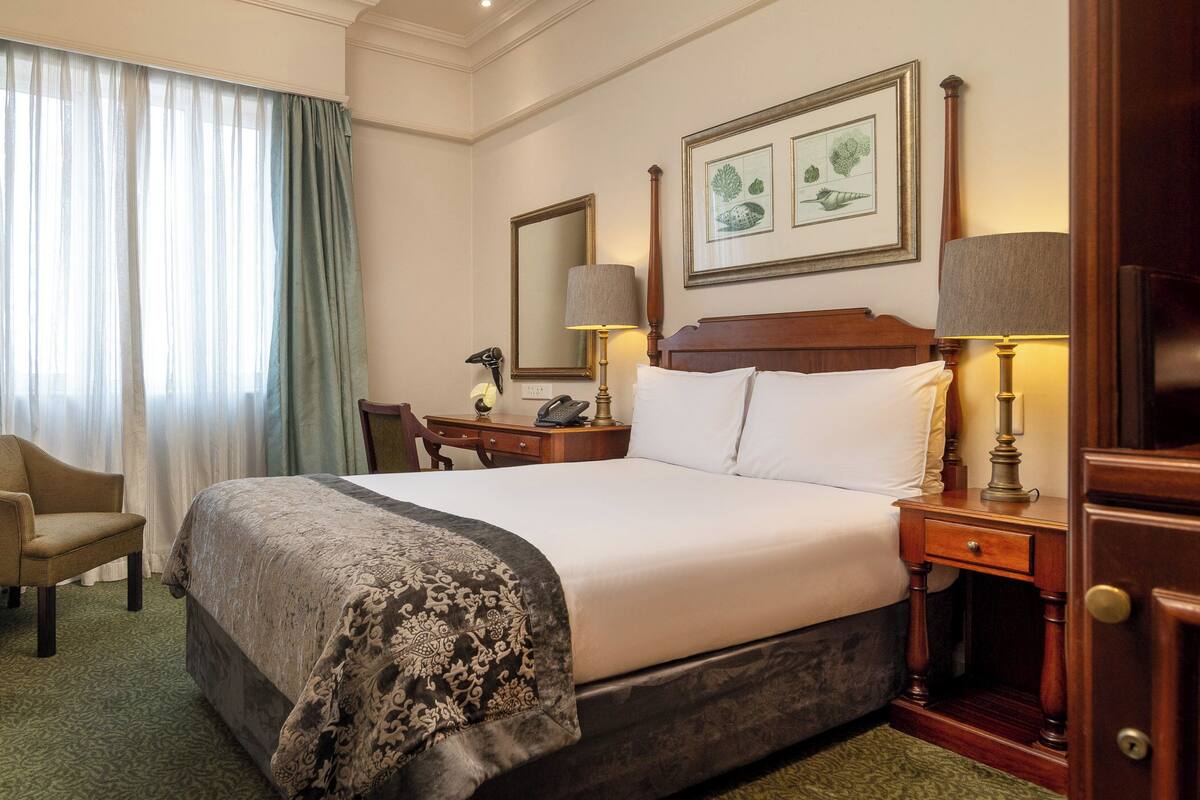Quarto de hotel com uma cama grande, uma poltrona, uma escrivaninha e uma mesa de cabeceira com luminárias.