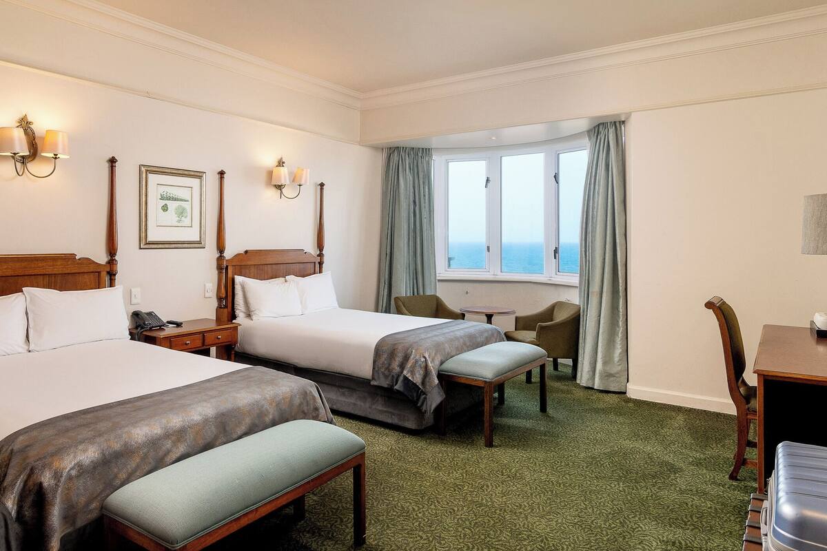 Quarto de hotel com duas camas, uma escrivaninha, uma cadeira e vista para o mar.