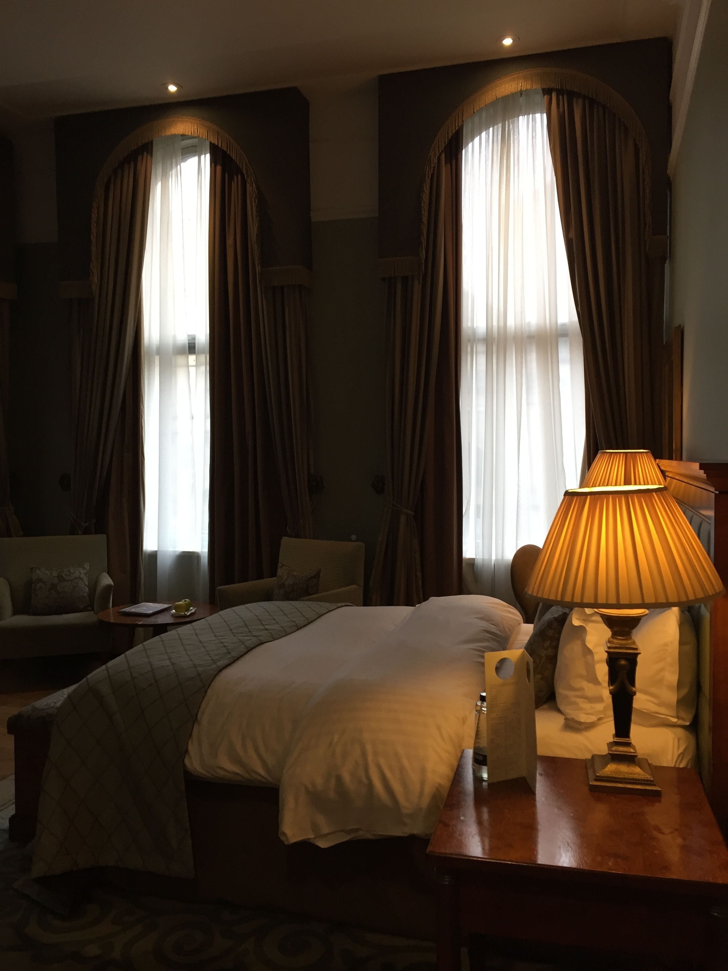 Quarto de hotel com uma cama, uma mesa de cabeceira com abajur, um sofá e duas janelas com cortinas.