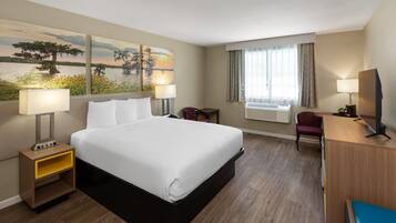 Quarto de hotel com uma cama grande, uma escrivaninha, uma cadeira, uma mesa de cabeceira, um abajur e uma pintura de um lago.