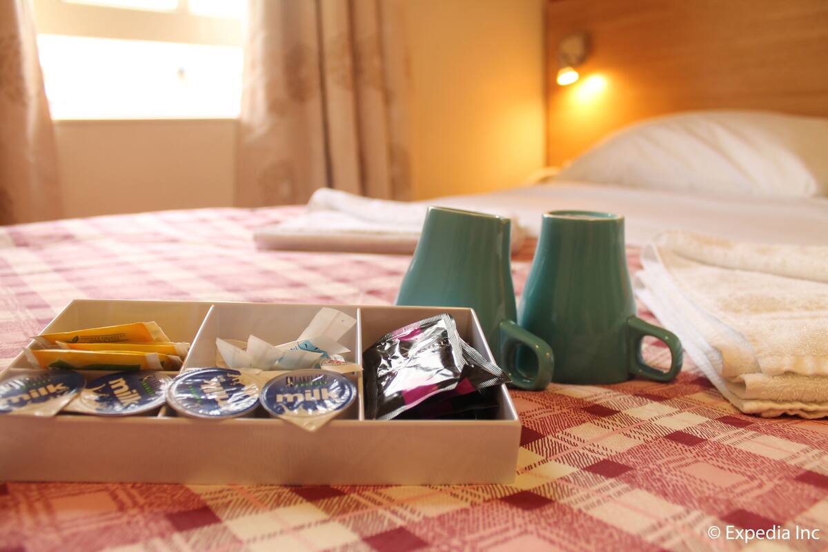 Cama bem arrumada com colcha xadrez, uma caneca verde e uma caixa de produtos de higiene pessoal sobre a mesa do quarto de hotel.