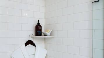 Uma casa de banho com paredes revestidas a azulejos brancos, cabine de duche em vidro, um roupão branco pendurado num gancho e uma prateleira com produtos de higiene pessoal.