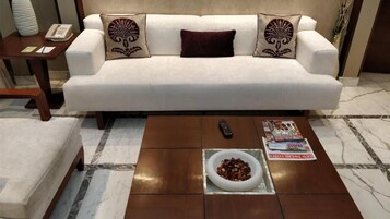 Um sofá branco com almofadas estampadas, uma mesa de centro de madeira com uma tigela de nozes e revistas.