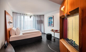 Quarto de hotel moderno com cama, escrivaninha com computador, mesinha com um vaso e uma janela grande com cortinas.
