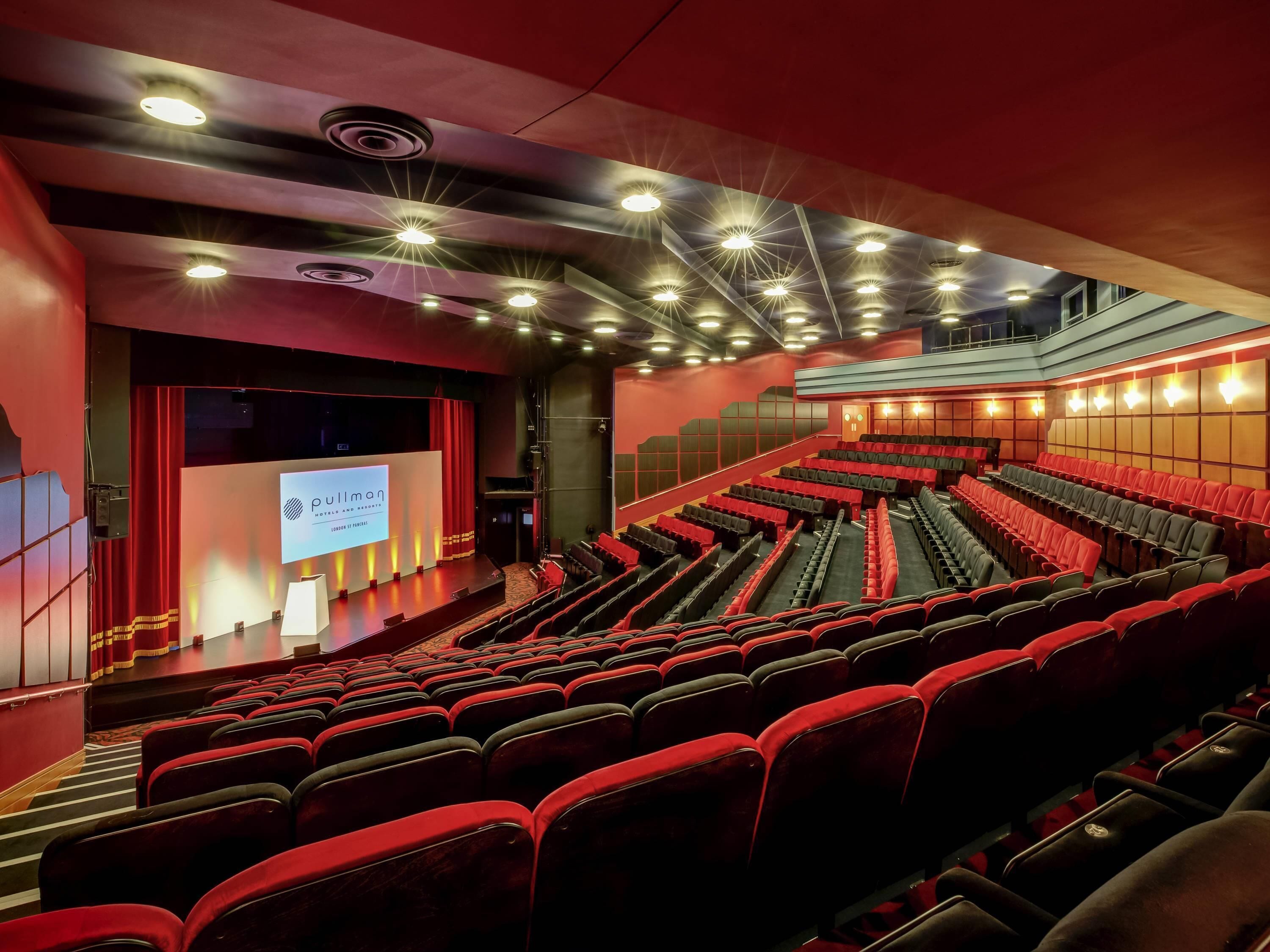 Um teatro com fileiras de assentos vermelhos e pretos, um palco com uma tela de projeção e uma área de balcão.