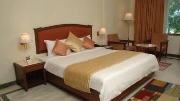 Quarto de hotel com uma cama grande, duas mesas de cabeceira, uma cadeira, uma mesinha e uma janela com cortinas.