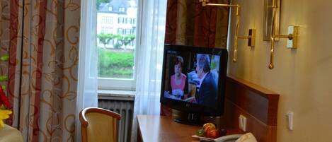 Quarto de hotel com televisão, uma mesa com controle remoto, uma cadeira e um sofá.