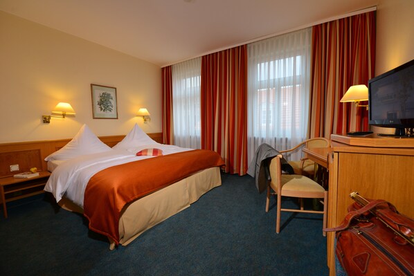 Quarto de hotel com cama, duas luminárias de cabeceira, televisão, escrivaninha com cadeira e mala.