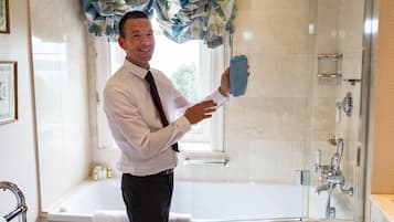 Um homem de camisa branca e gravata escura segurando uma toalha azul num banheiro com box de vidro, uma banheira branca e uma janela com cortinas.
