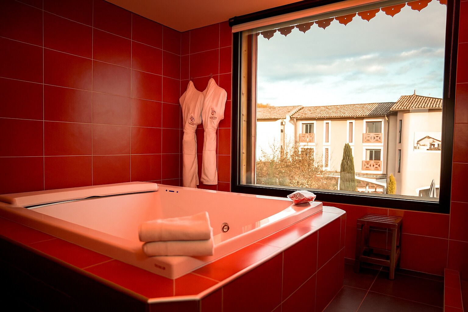 Um banheiro com azulejos vermelhos, uma banheira grande, uma janela com vista para edifícios e um banquinho pequeno.