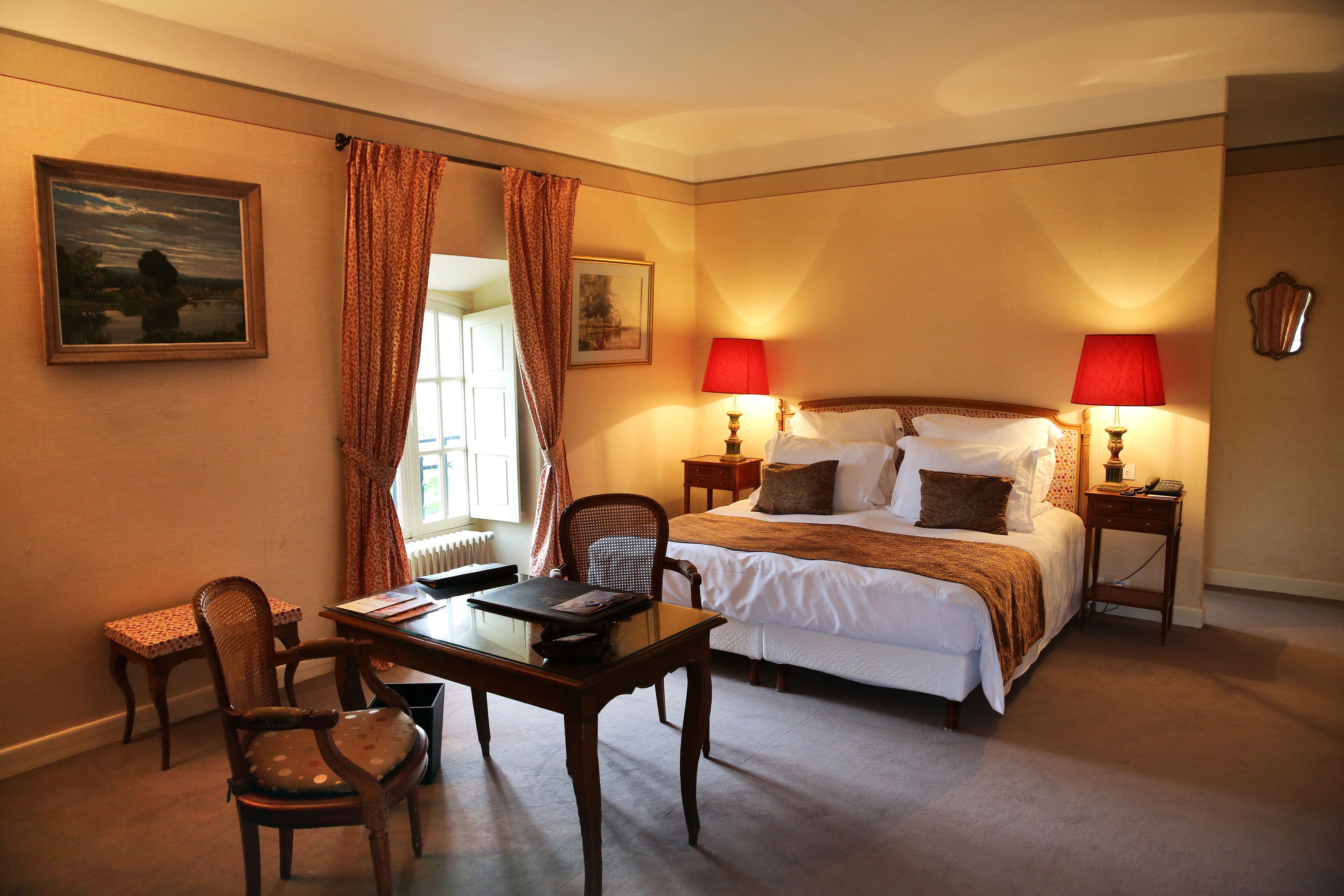 Quarto de hotel com uma cama, duas luminárias de cabeceira, uma escrivaninha com cadeira, uma mesa pequena e uma janela com cortinas.