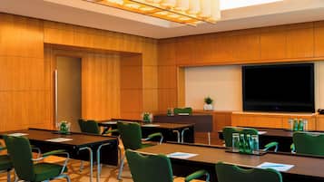 Sala de conferências com painéis de madeira, uma grande TV de tela plana, cadeiras verdes e mesas com garrafas de água.
