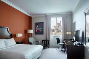 Quarto de hotel com uma cama grande, uma escrivaninha, uma cadeira e vista para um edifício histórico.