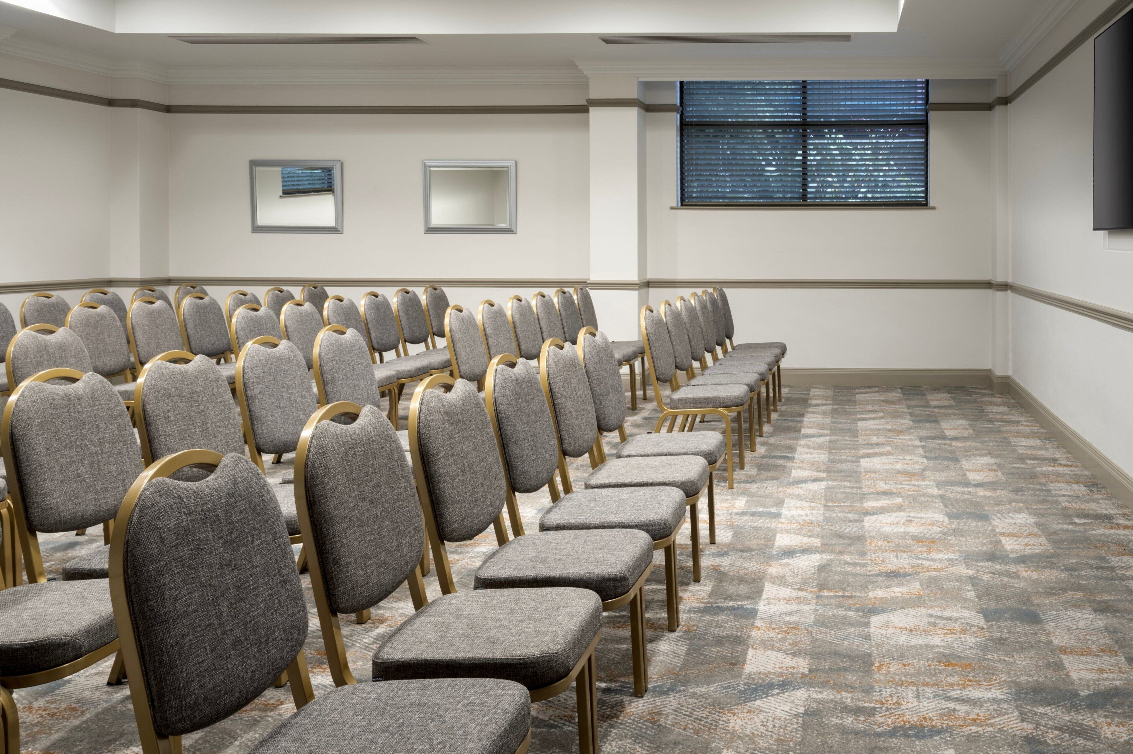 Sala de conferências com fileiras de cadeiras cinzas e um tapete estampado.