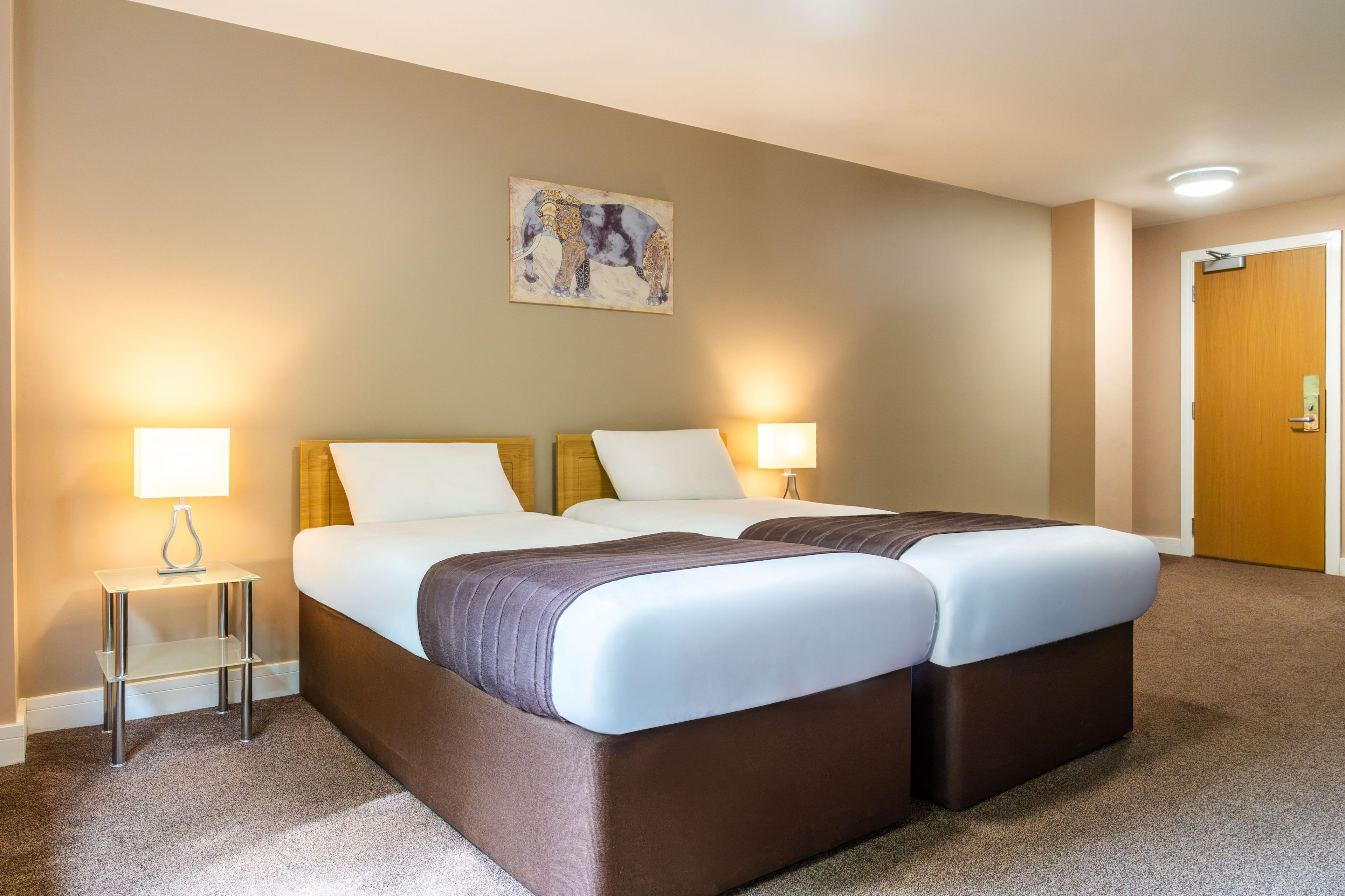 Quarto de hotel com cama de casal, duas mesas de cabeceira, um criado-mudo, um quadro na parede e uma porta.