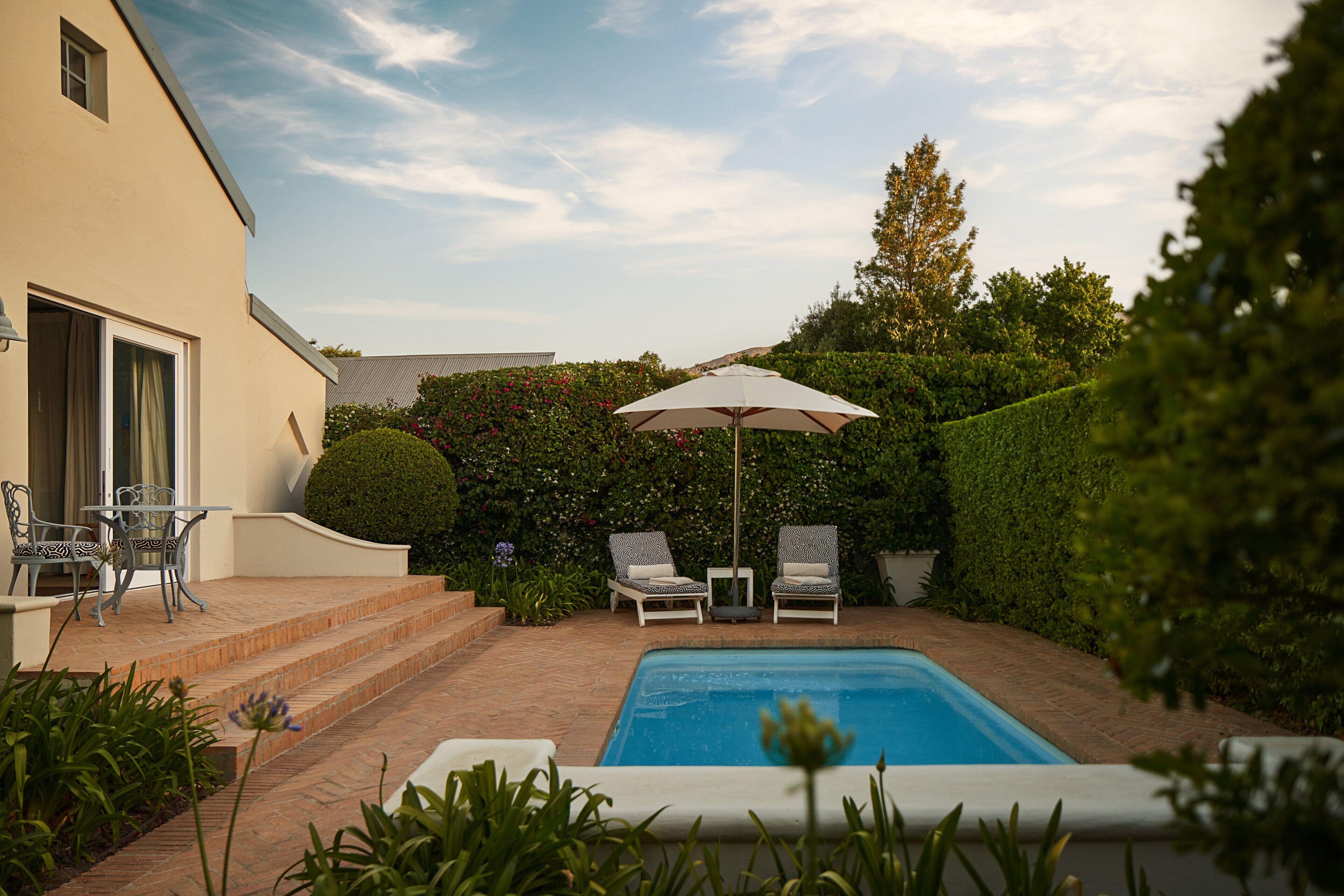 Piscina com área de terraço, incluindo mesa e cadeiras, cercada por vegetação e um hedge.