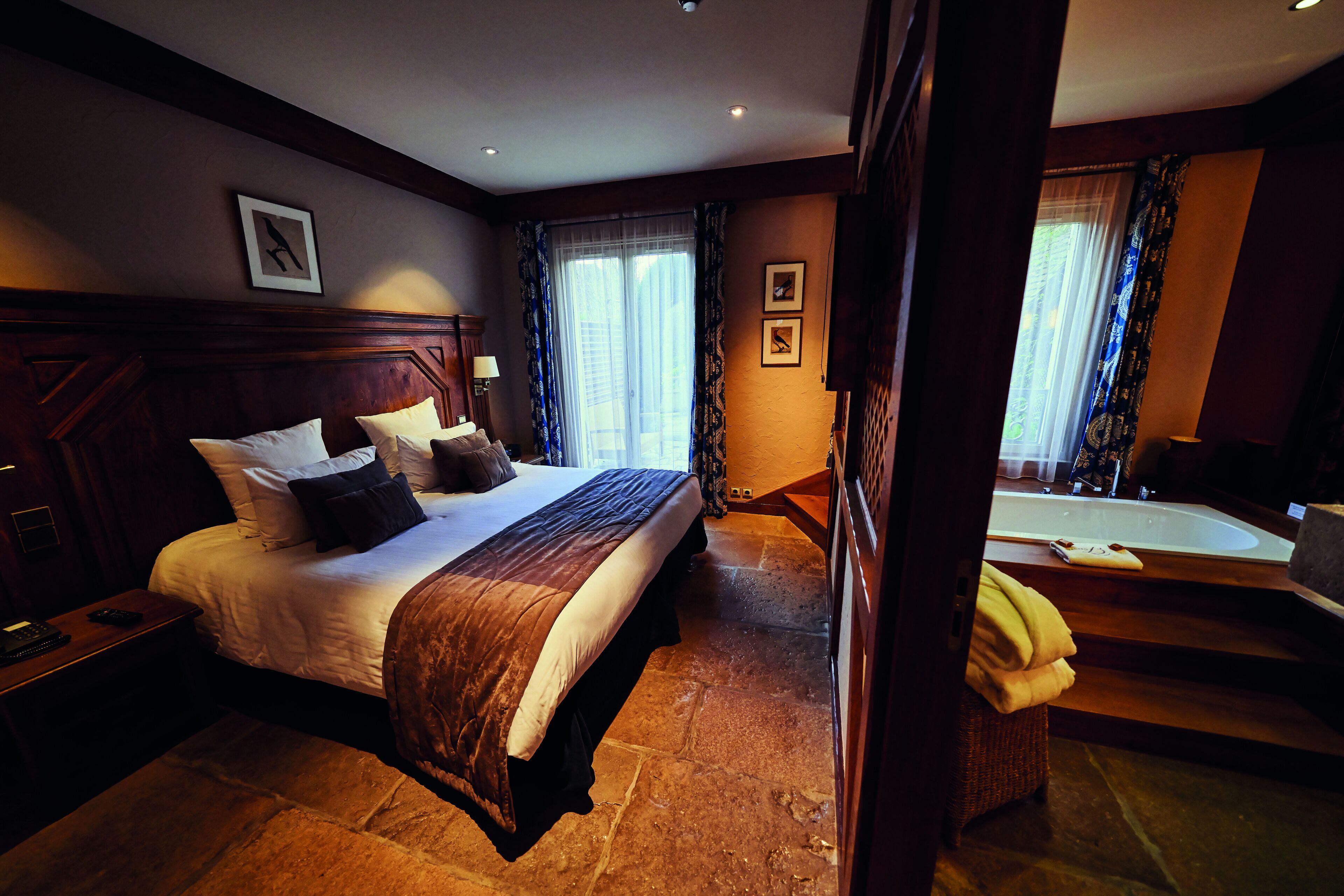 Quarto de hotel com uma cama grande, cabeceira de madeira, mesa de cabeceira, janelas com cortinas, banheiro com banheira e piso de madeira.