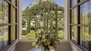Quarto com vista para uma paisagem montanhosa, um vaso com flores sobre uma mesa e uma janela com cortinas.