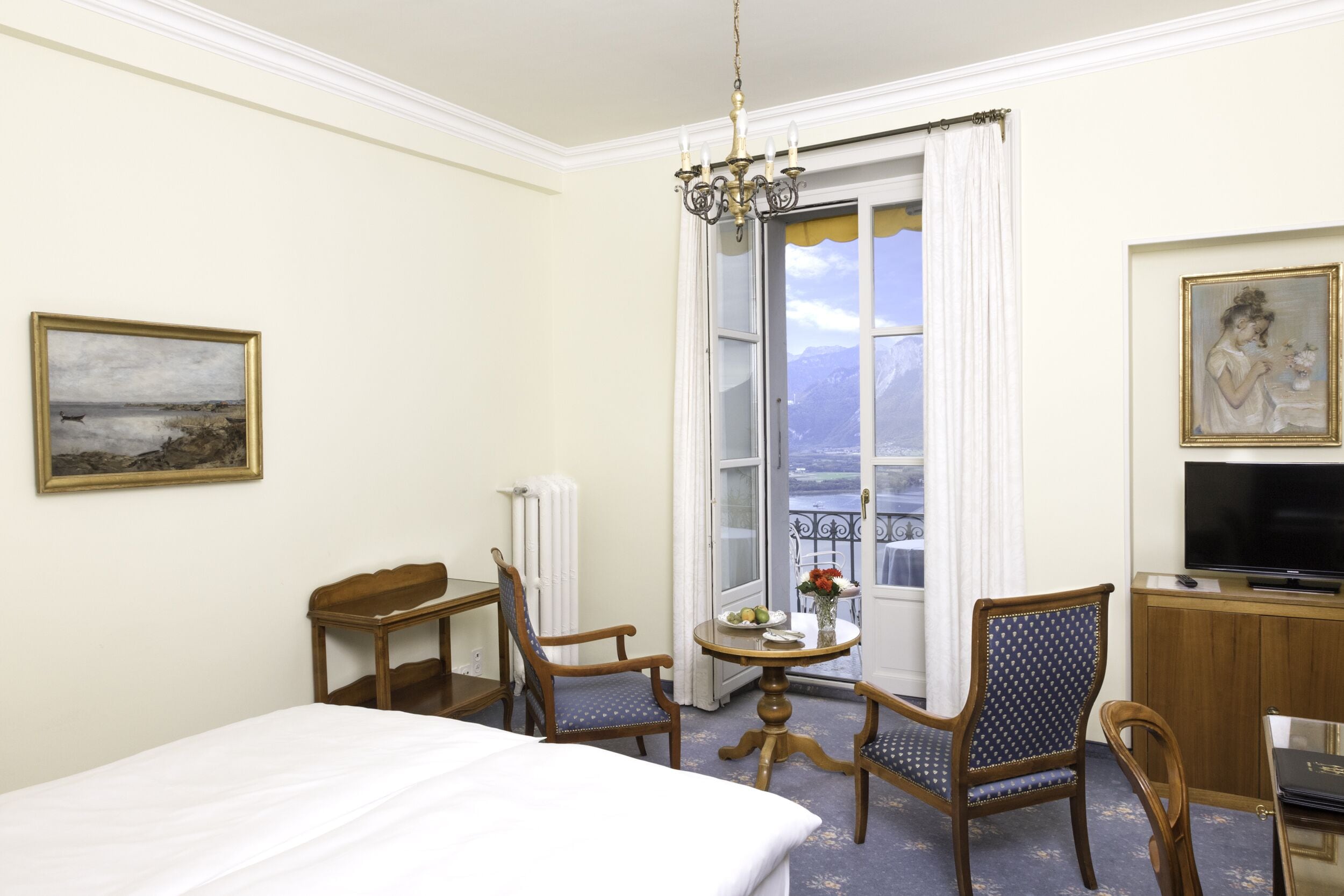 Quarto de hotel com cama, duas cadeiras, uma mesinha com um vaso, televisão e um quadro na parede.