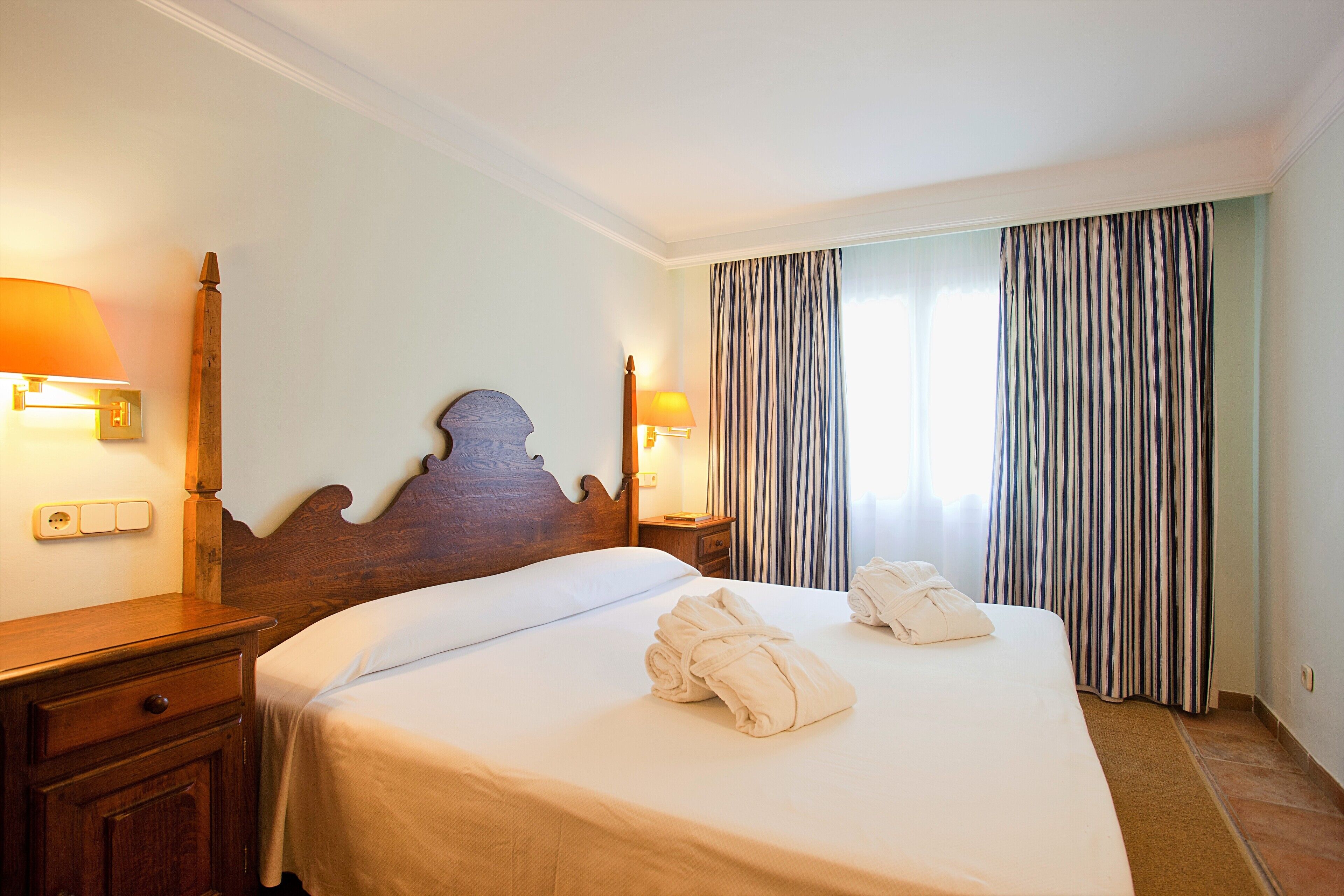 Quarto de hotel com cabeceira de madeira, cama com lençóis brancos, duas luminárias de cabeceira, mesa de cabeceira de madeira e uma janela com cortinas listradas.
