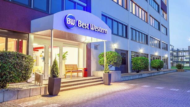 Entrada de hotel com uma placa indicando "BW Best Western". Há degraus que levam à entrada, um banco e vasos de plantas.