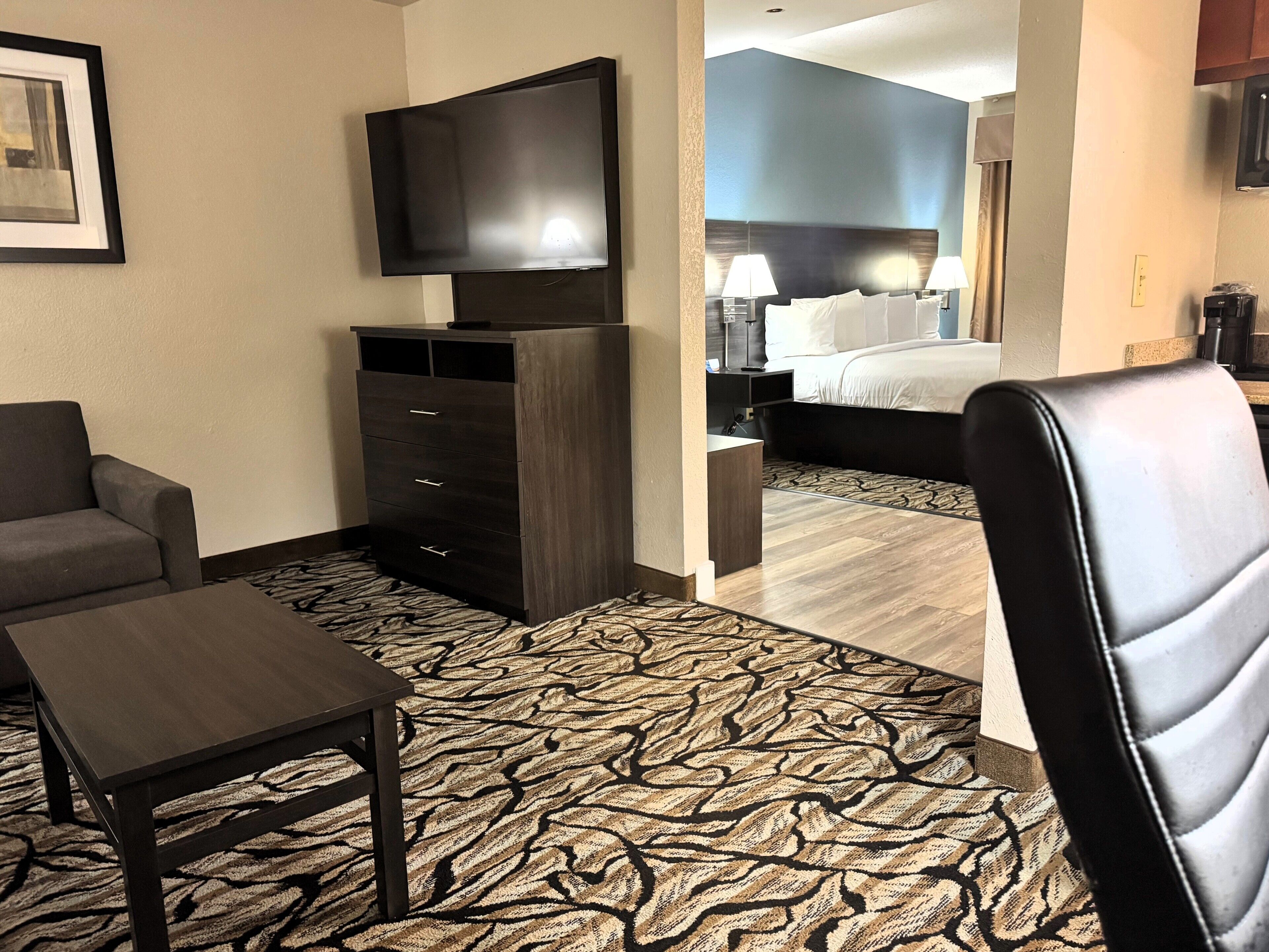 Quarto de hotel com televisão de tela plana na parede, cómoda de madeira escura, carpete estampado, poltrona de couro e cama com roupa de cama branca.