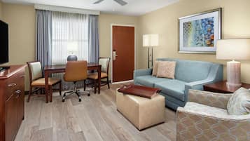 Quarto de hotel com televisão, mesa de jantar com cadeiras, sofá, poltrona e luminária.