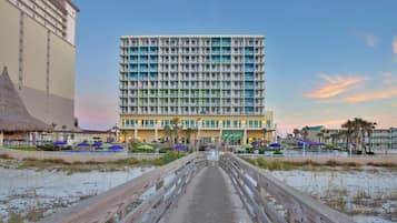 Um hotel à beira-mar com um pátio central, uma passarela que leva à entrada e uma estrutura com telhado de palha à esquerda.