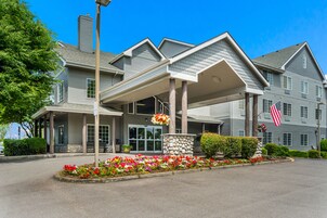Um hotel de dois andares com entrada coberta, um pilar de pedra e um canteiro de flores na frente.