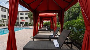 Uma área de estar ao ar livre coberta, com toldos vermelhos, espreguiçadeiras à beira da piscina e uma piscina.