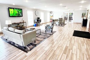 Uma sala de estar moderna com sofá, mesa de centro e televisão. Há uma escrivaninha com computador e uma cadeira. O chão é de madeira e possui um tapete estampado.