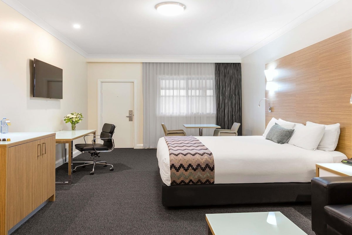 Quarto de hotel com cama, escrivaninha, cadeira e televisão na parede.