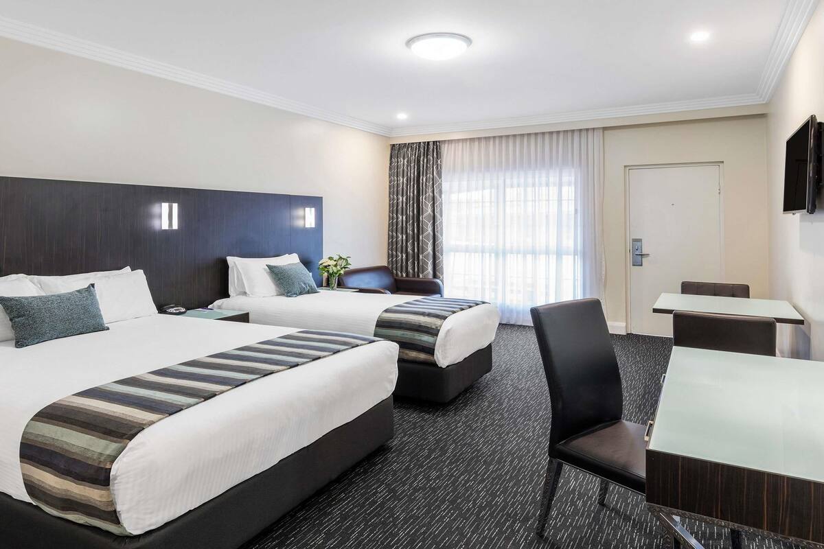 Quarto de hotel com duas camas, uma escrivaninha, uma cadeira, uma TV e uma janela com cortinas.