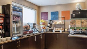 Espaço de café da manhã de hotel, com máquina de bebidas automática, canto de café e diversas opções de alimentos à mostra.