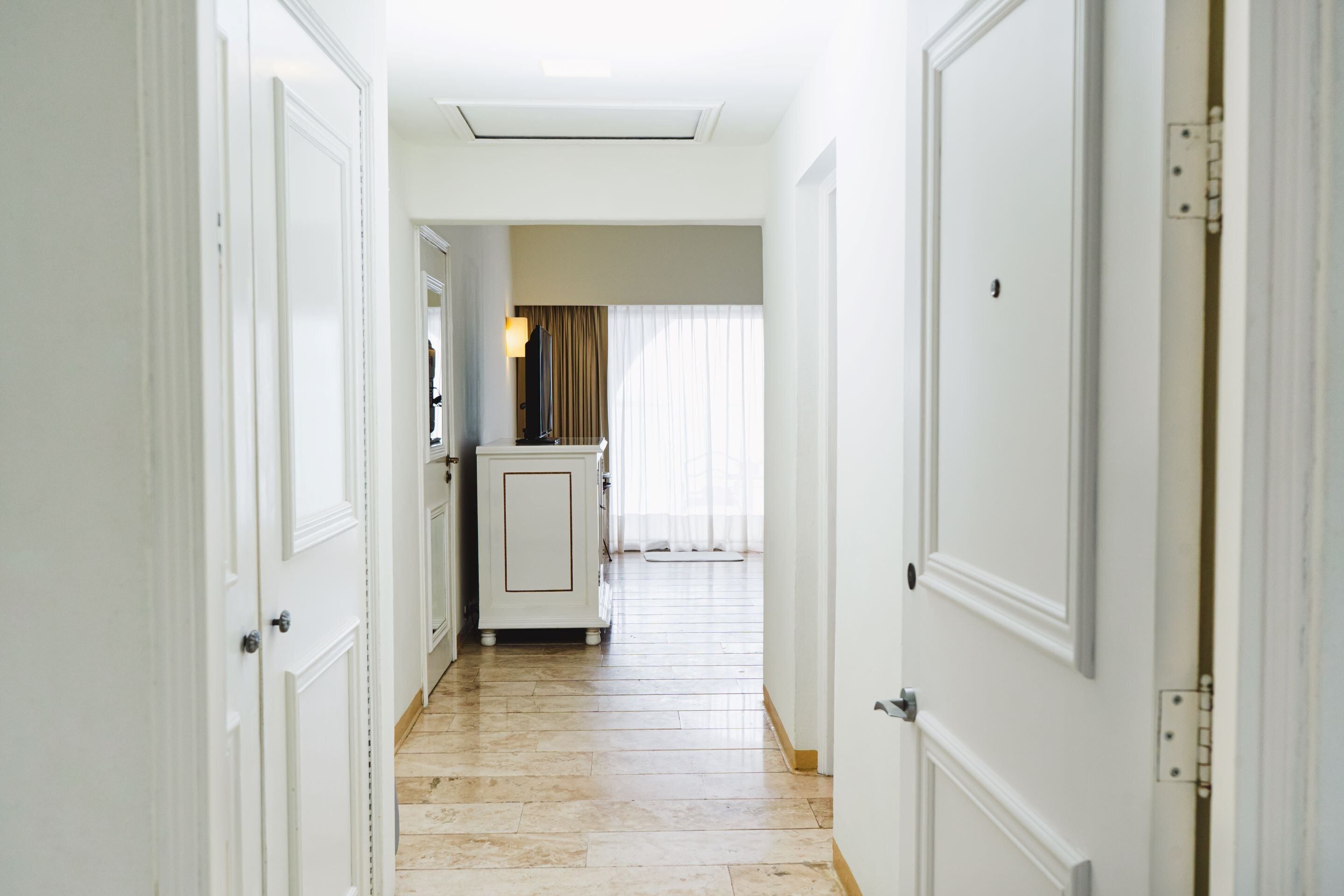 Um corredor com portas brancas, uma área de cozinha e vista para um quarto com janela e cortinas.