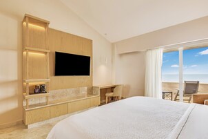 Quarto de hotel com uma cama grande, uma escrivaninha com cadeira, uma TV fixada na parede e uma varanda com vista para o mar.